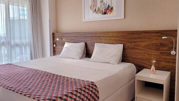 Quarto de hotel com uma cama grande, cabeceira de madeira, mesa de cabeceira com um vaso e uma moldura na parede.
