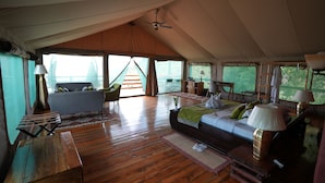 Acolhimento em tenda espaçosa com soalho de madeira, uma cama com almofadas, um sofá, uma cadeira e uma escrivaninha.
