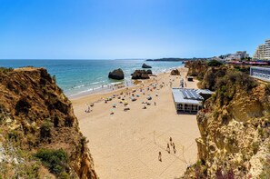 Uma praia com área arenosa, falésias rochosas e um edifício com telhado.