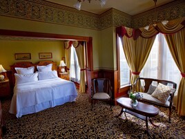 Quarto de hotel com uma cama grande, uma cadeira, uma mesinha e uma janela com cortinas.