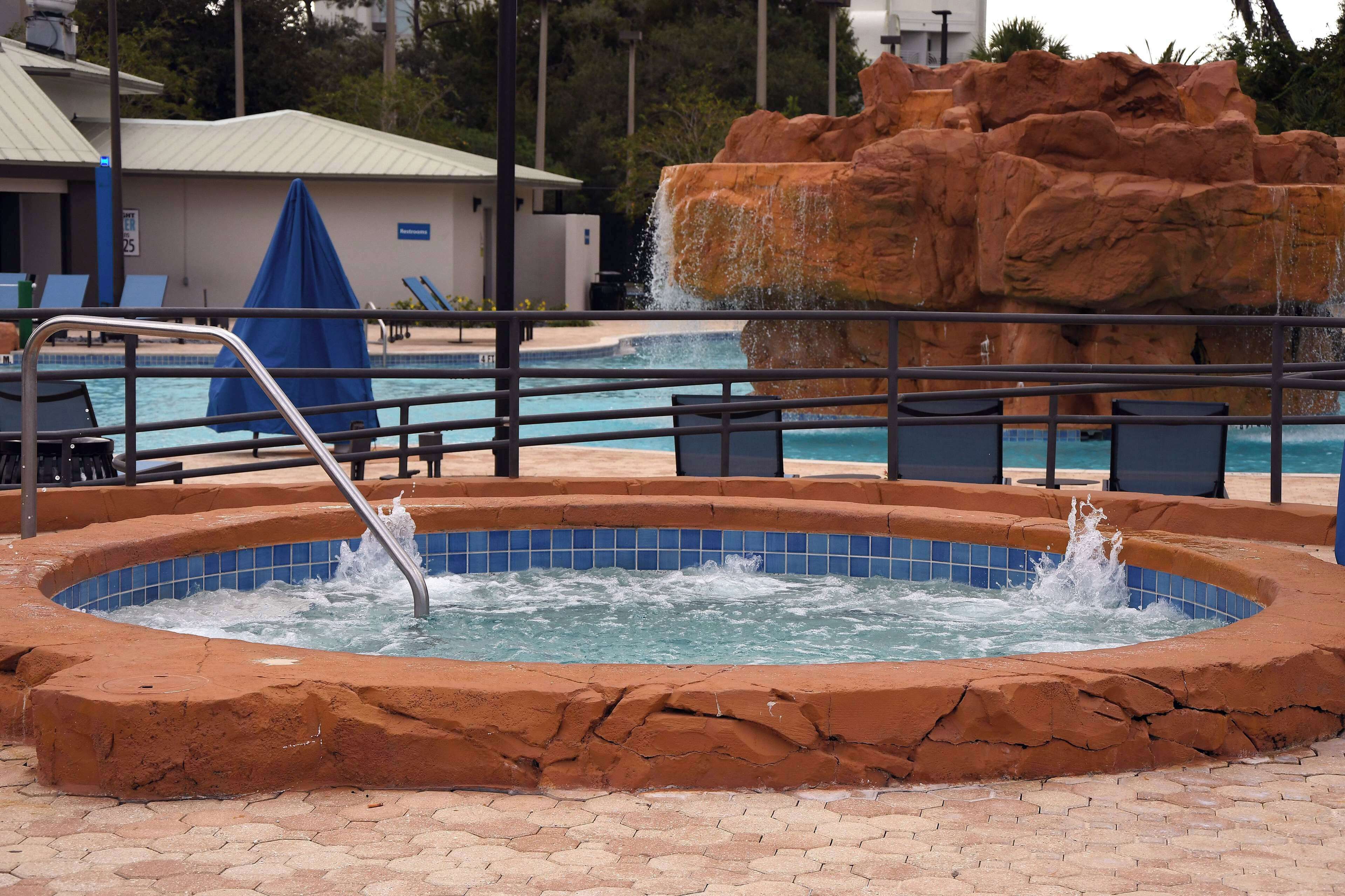 Uma banheira de hidromassagem com um cenário rochoso, rodeada por uma área de piscina com azulejos e espreguiçadeiras.