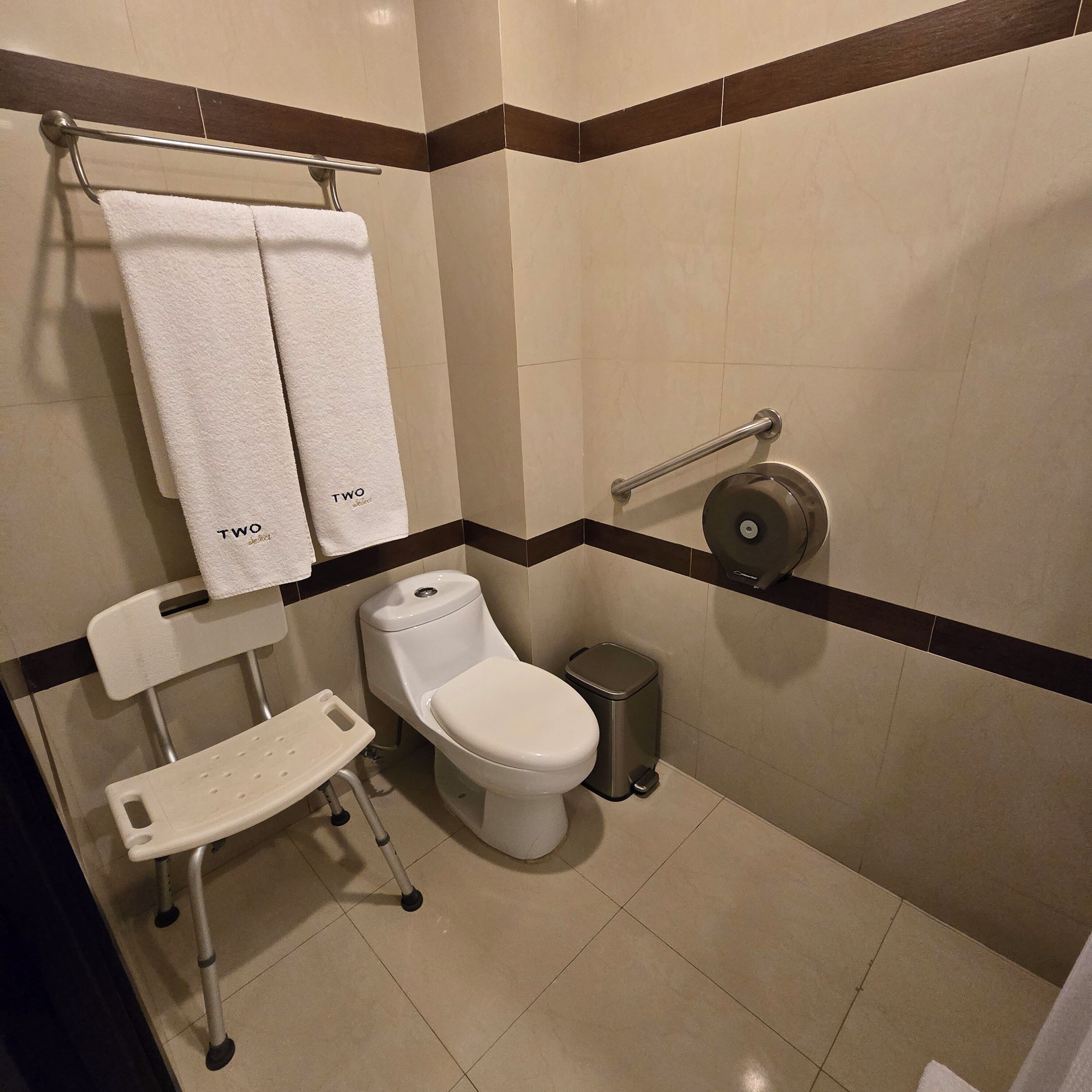 Um banheiro pequeno e moderno, com vaso sanitário, bidê e barra de apoio. Duas toalhas brancas penduradas em um suporte.