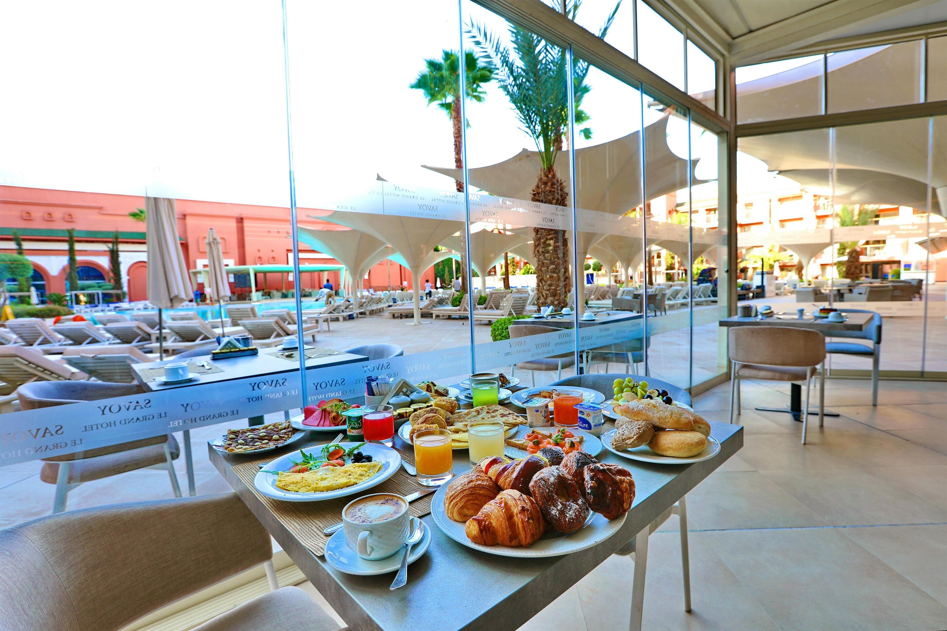 Um buffet de café da manhã com diversas opções de comida e bebidas, área de piscina com espreguiçadeiras e vista para um edifício com arcos.