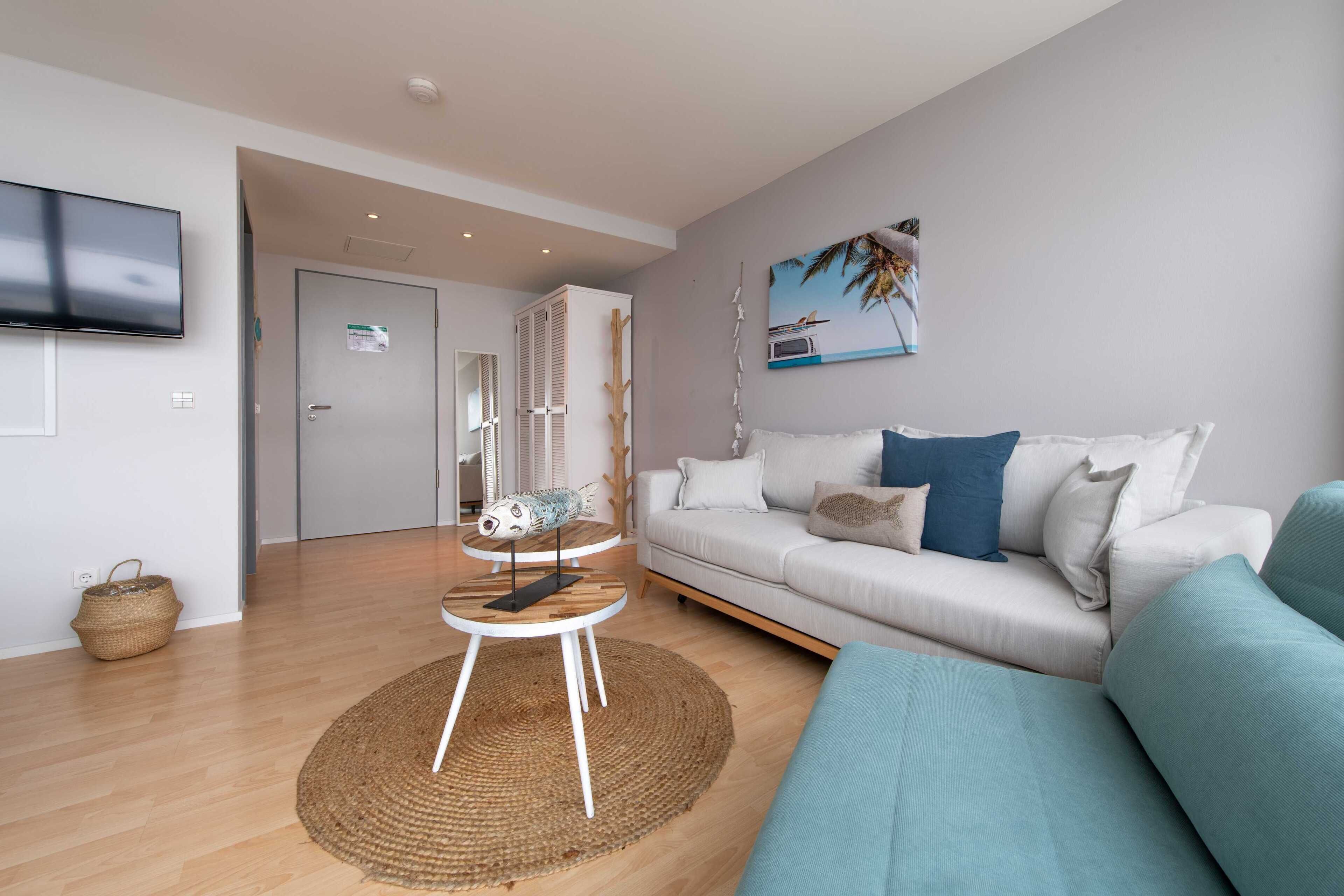 Uma sala de estar moderna com um sofá branco, uma poltrona azul, uma televisão de tela plana na parede e piso de madeira.