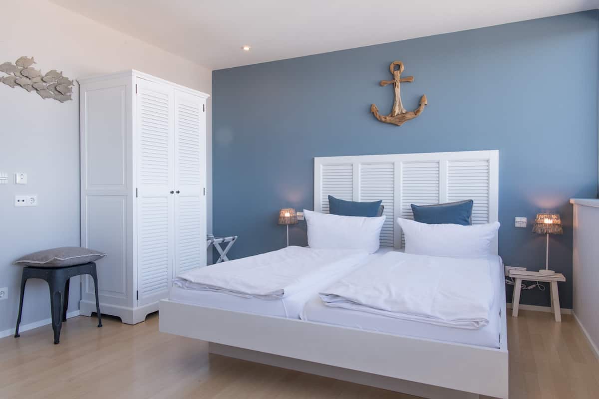 Quarto com cama, cabeceira branca, duas mesas de cabeceira com abajures, um criado-mudo, um banquinho pequeno e um guarda-roupa.