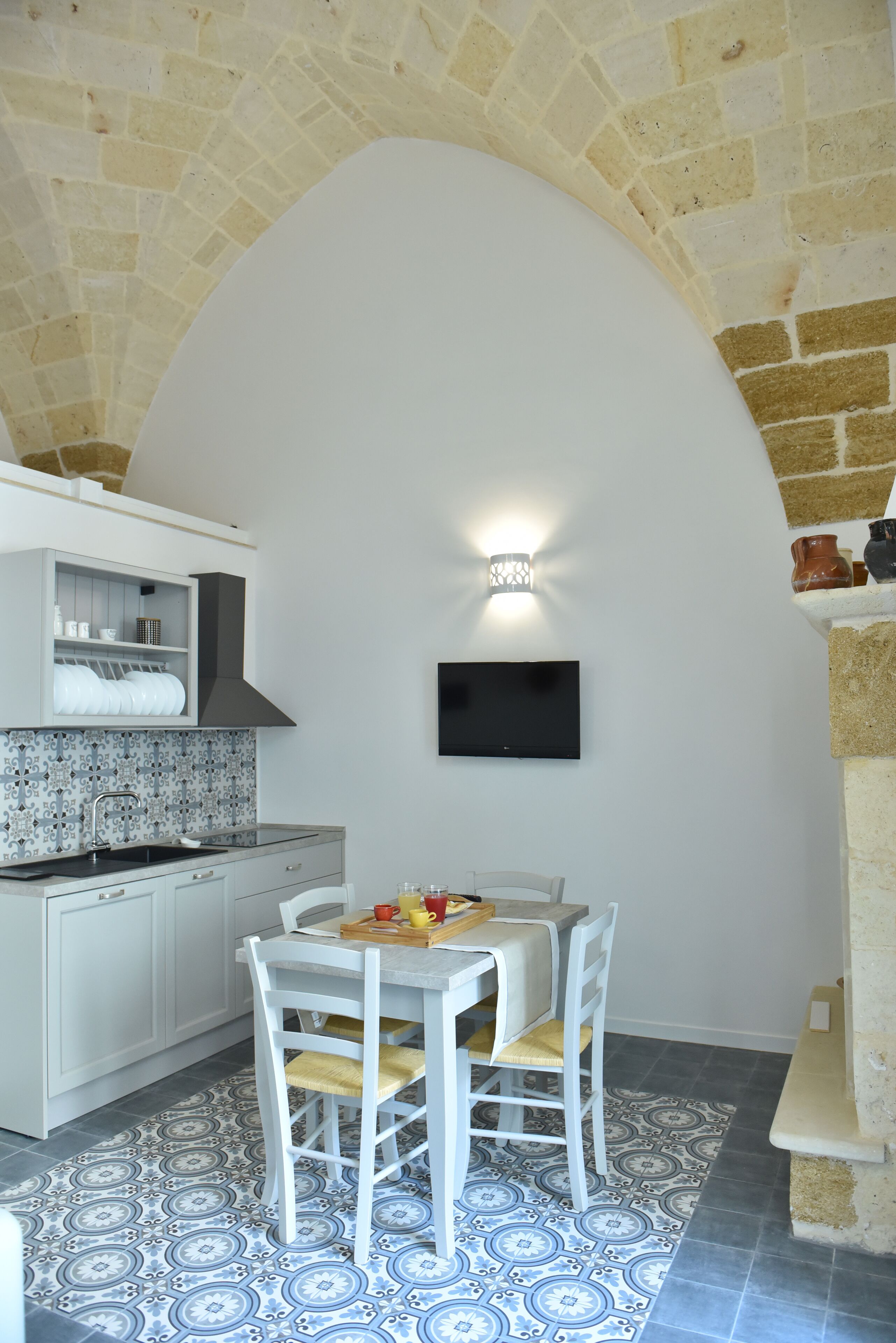 Uma cozinha com uma mesa e cadeiras brancas, um revestimento de azulejos e um arco de pedra.