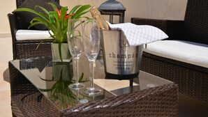 Uma cadeira de vime com almofada, uma mesa de vidro com uma garrafa de champanhe, dois copos de vinho e um vaso de planta.