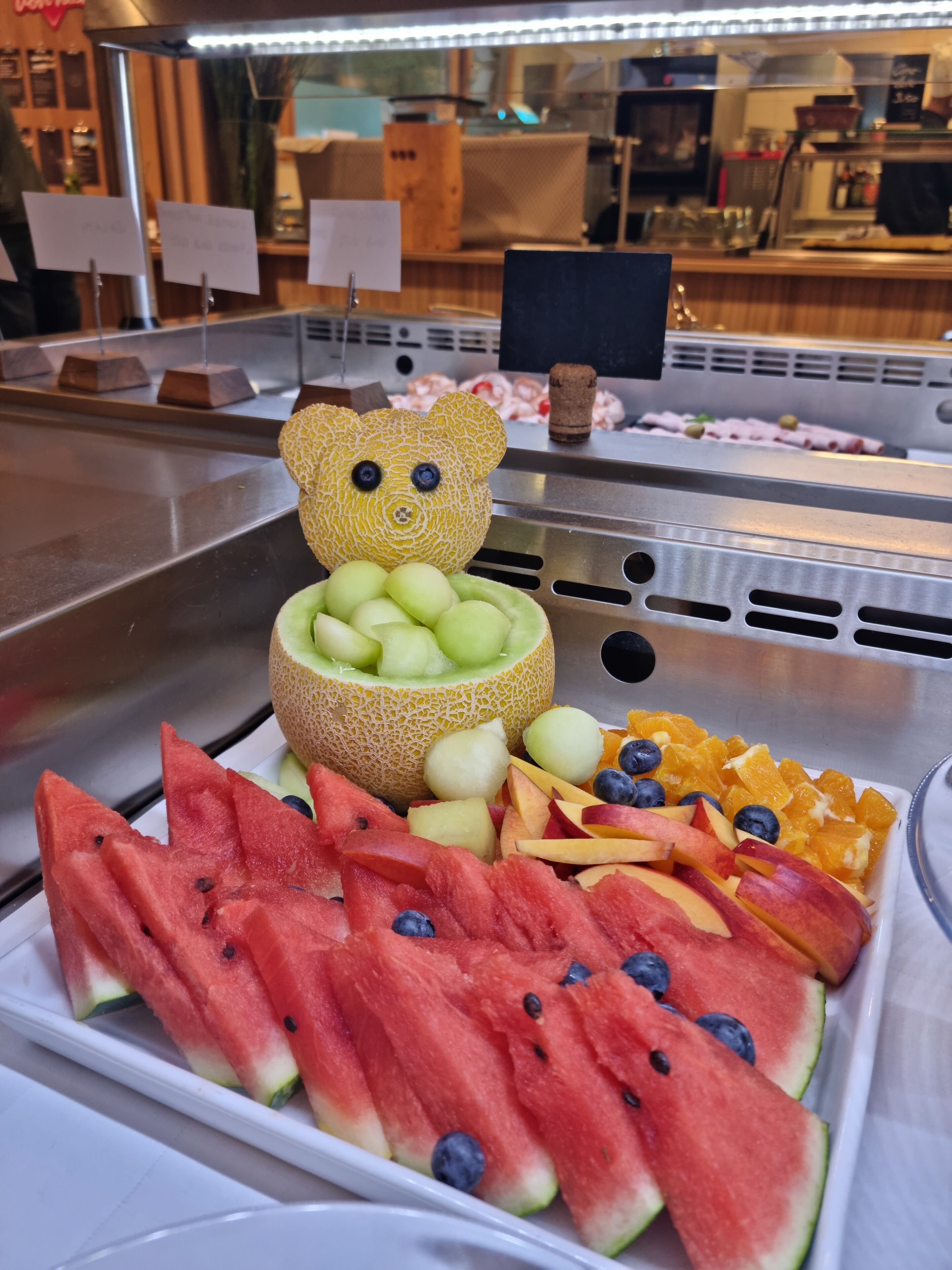 Exposição de frutas com uma tigela de melão em formato de favo de mel, fatias de melancia e diversas frutas.