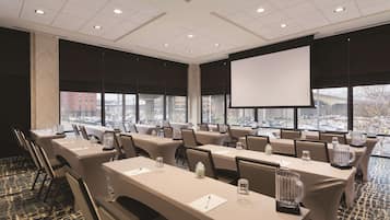 Sala de conferências com mesas, cadeiras, tela de projetor e amplas janelas com vista para edifícios e estacionamento.