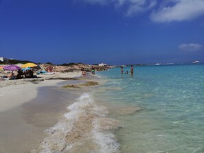 Uma praia com águas turquesa cristalinas, pessoas tomando sol e um píer ao longe.