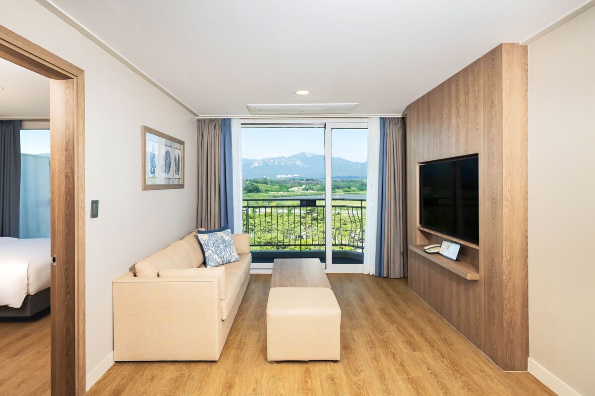 Quarto de hotel com sofá, pufê, televisão e varanda com vista para as montanhas.