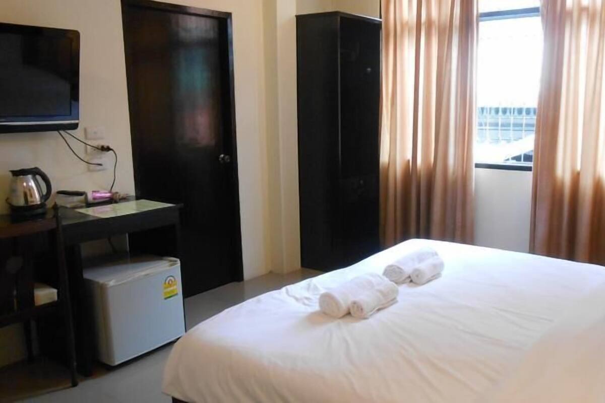 Quarto de hotel com cama, escrivaninha com chaleira, guarda-roupa e janela com cortinas.