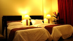 Quarto de hotel com duas camas, cada uma com um cobre-leito branco e um cobertor vermelho. Há duas luminárias de mesa e um vaso de planta do lado direito.