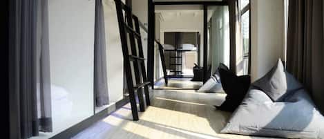 Corredor moderno de hotel com piso de madeira, escada para uma cama beliche e uma janela grande que permite entrada de luz natural.