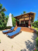 Resort à beira-mar com área de estar coberta, duas espreguiçadeiras com guarda-sóis e uma escadaria para a entrada.