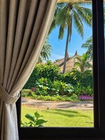 Janela com cortinas, vista de um jardim tropical com palmeiras, uma construção com telhado de palha e diversas plantas e flores.