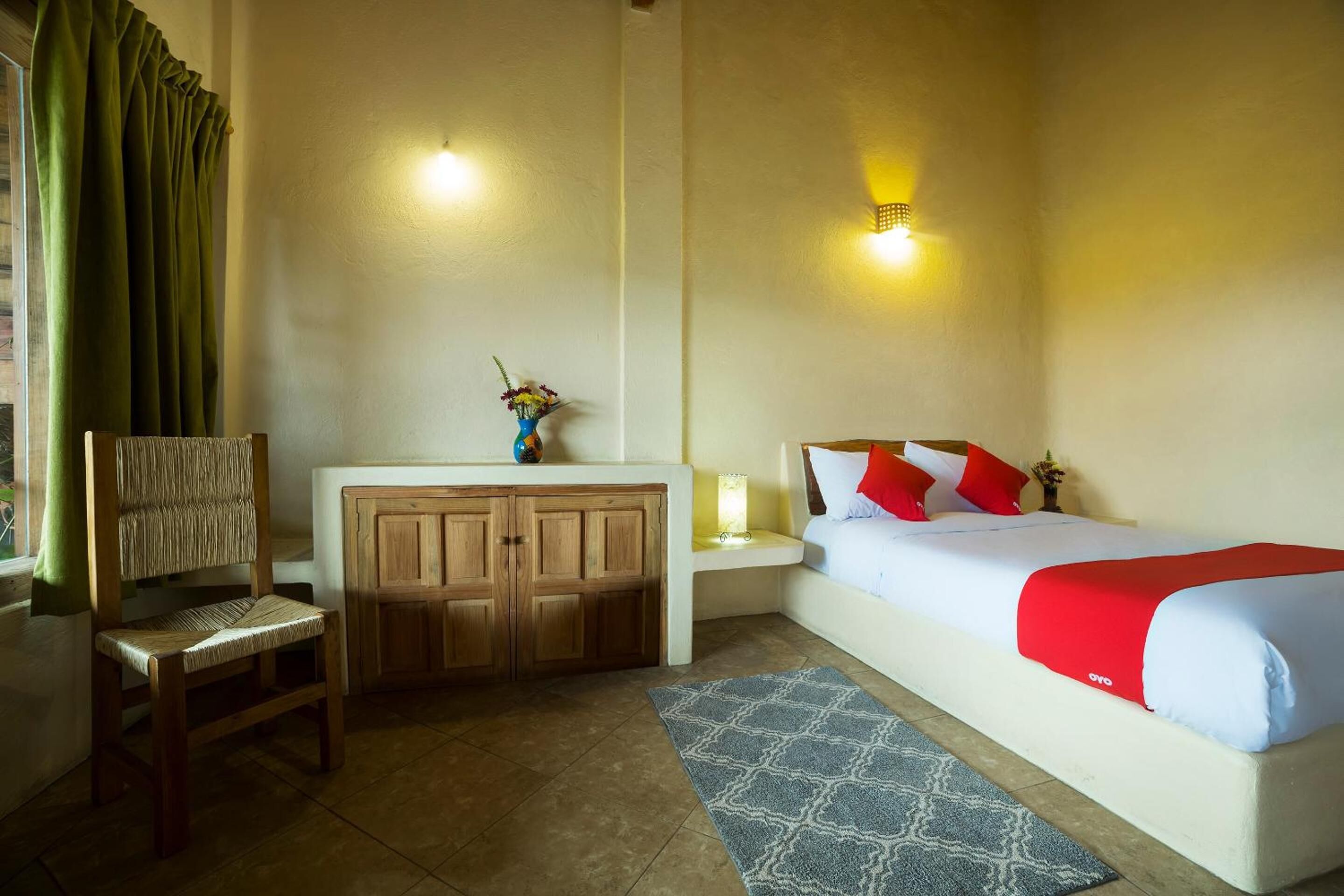 Quarto de hotel com cama, mesa de cabeceira de madeira, uma cadeira, uma mesinha com um vaso e luminária na parede.