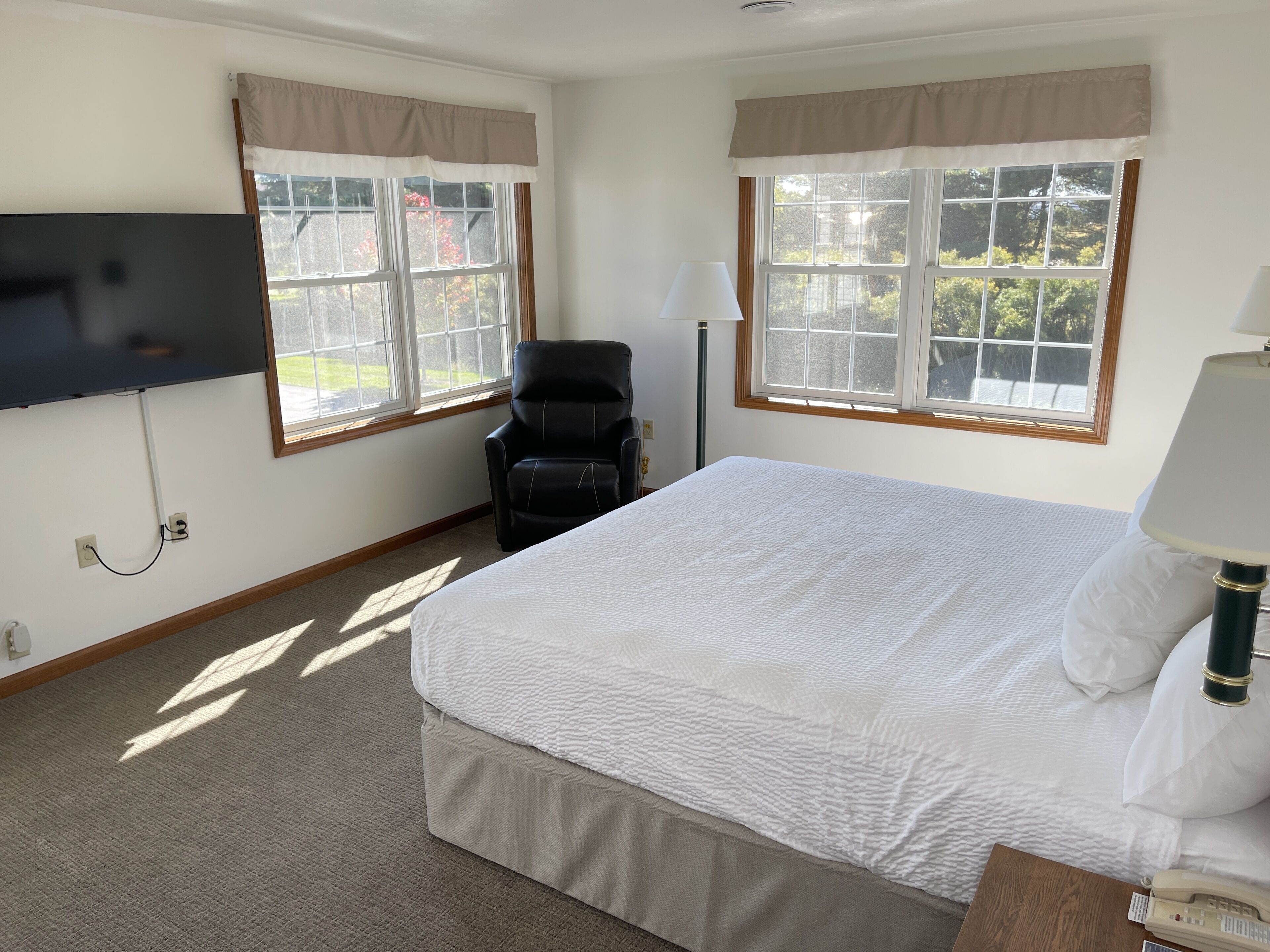 Quarto de hotel com uma cama grande, duas janelas com cortinas, uma cadeira preta, uma escrivaninha com luminária e uma televisão fixada na parede.