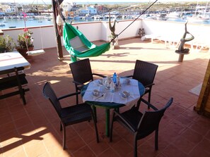 Terraço com mesa redonda posta para quatro pessoas, rodeado de cadeiras, com vista para uma marina com barcos.