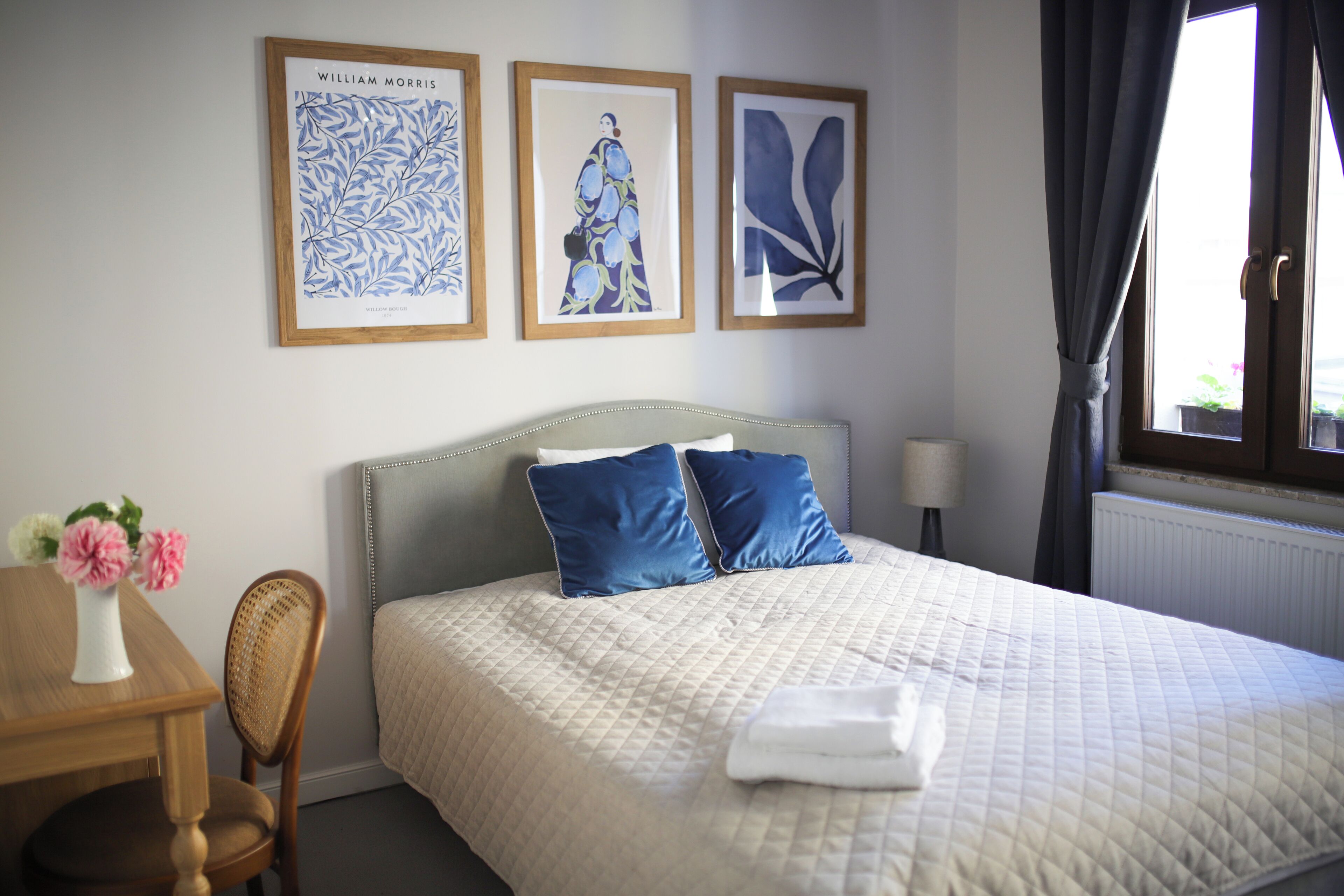 Quarto de hotel com uma cama, uma cadeira, uma mesa com um vaso de flores e três obras de arte emolduradas na parede.