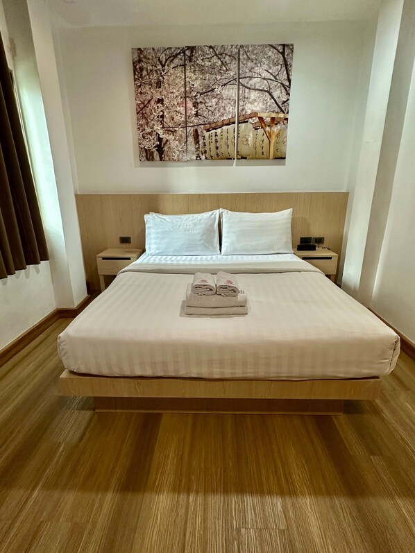 Cama bem arrumada com lençóis brancos e toalha dobrada, cabeceira de madeira, mesa de cabeceira com abajur e arte na parede com cerejeiras.