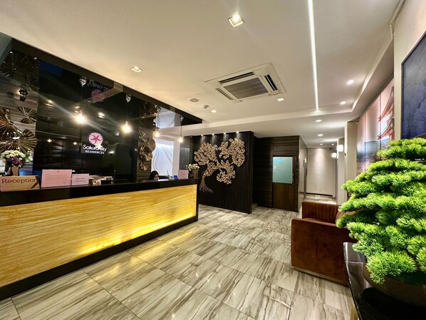 Uma área de recepção de hotel, com balcão, tela digital e placa indicativa.