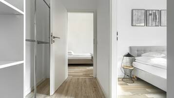 Quarto com uma cama, mesa de cabeceira e criado-mudo. Há uma porta que dá para outro cômodo.