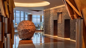 Um lobby moderno com uma grande escultura de madeira, uma parede de pedra com padrão geométrico e vista para a cidade através de amplas janelas.