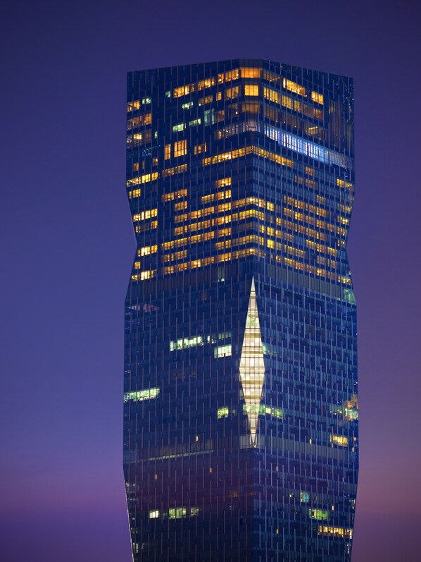 Um edifício moderno de grande altura com fachada de vidro triangular marcante e inúmeras janelas iluminadas à noite.