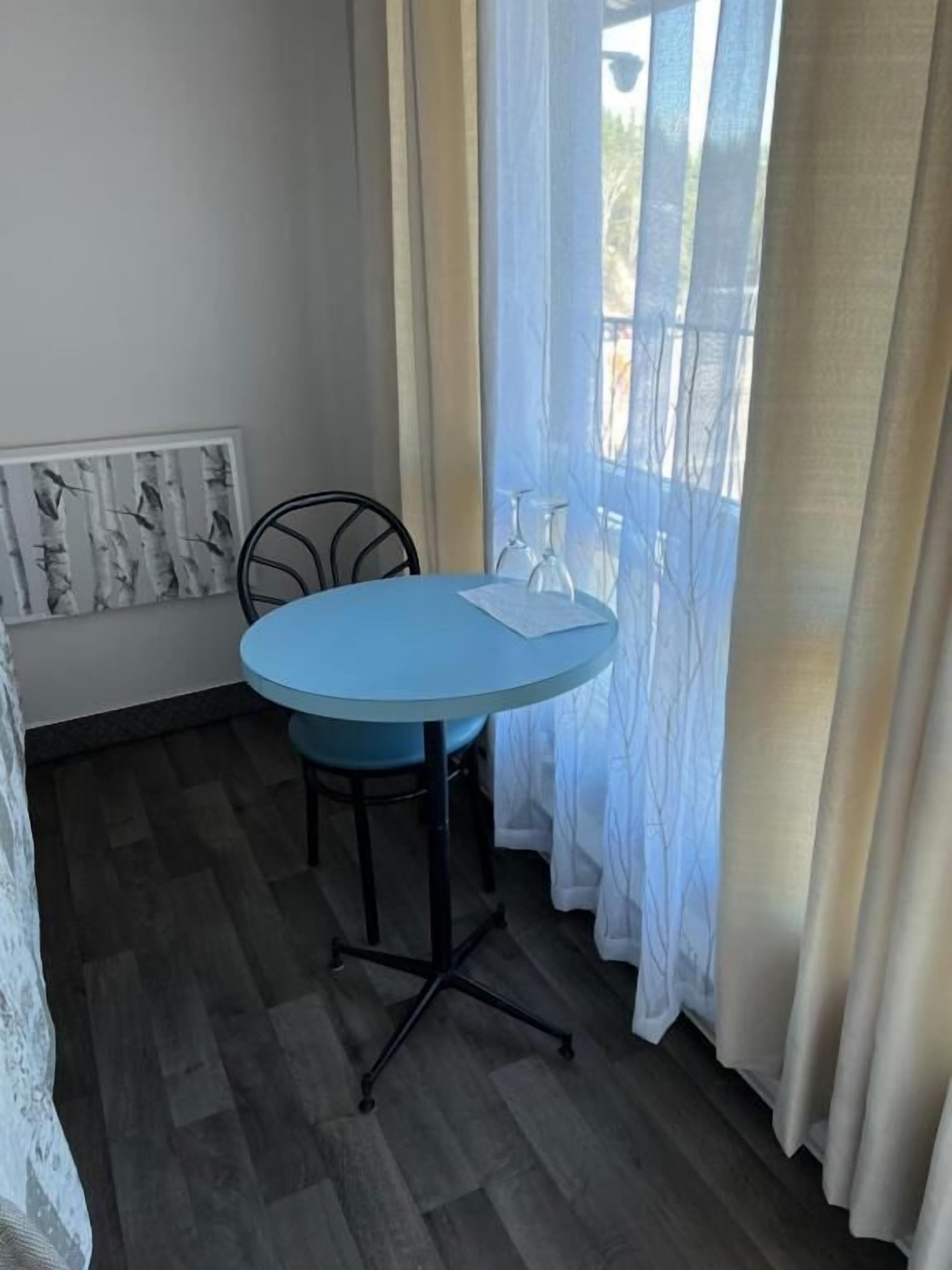 Quarto pequeno com uma mesa redonda e duas cadeiras, uma janela com cortinas leves e parede com padrão.
