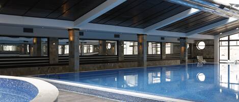 Piscina interna com borda azulejada, área de estar com cadeiras e teto alto com dutos aparentes.