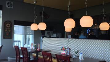 Interior de restaurante com mesas e cadeiras de madeira, luminárias pendentes e um suporte para o menu.