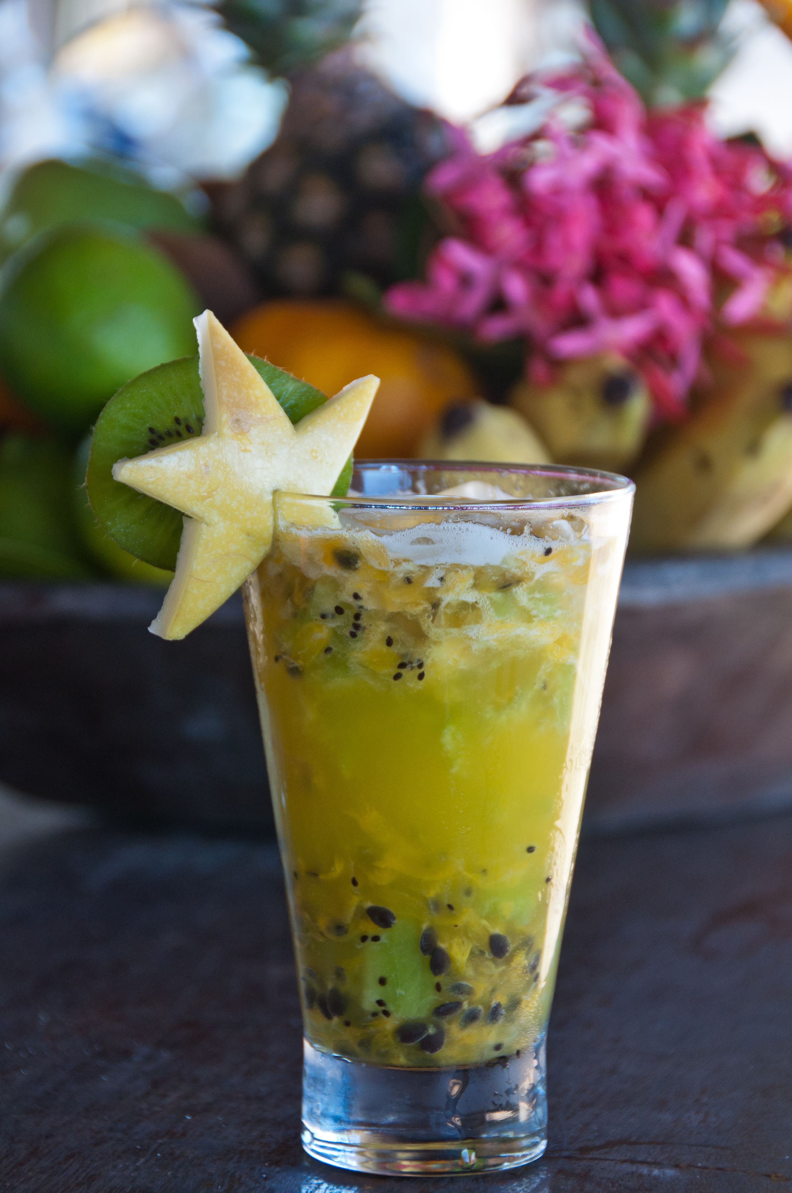 Um copo alto com um enfeite em formato de estrela, cheio de uma bebida amarela e rodeado por frutas.