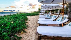 Uma área de lounge à beira-mar com espreguiçadeiras e guarda-sóis, um caminho de areia e vista para o oceano.