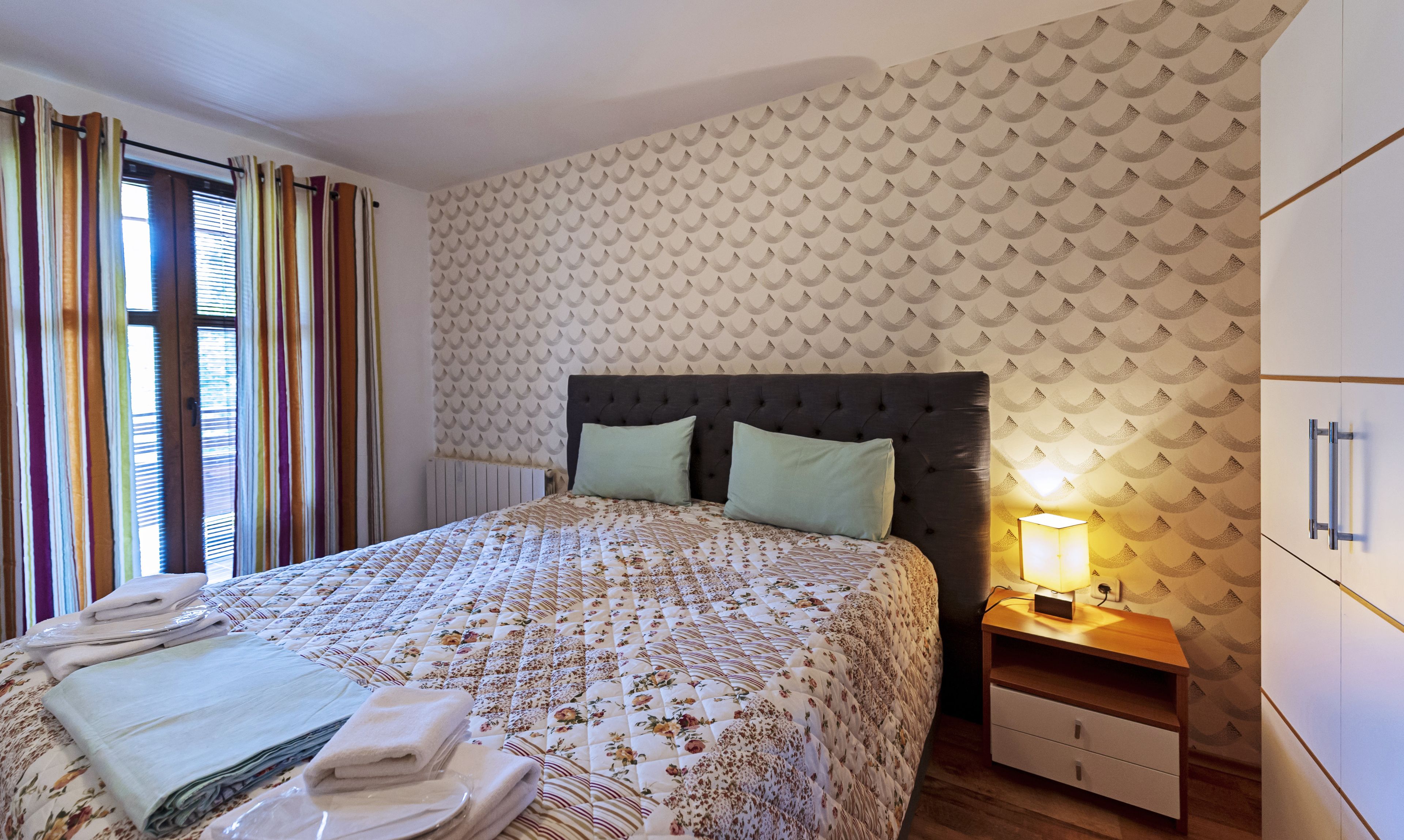 Cama bem arrumada com colcha florida, mesa de cabeceira com abajur e guarda-roupa.