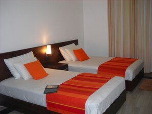 Quarto de hotel com duas camas, cada uma com roupa de cama branca e almofadas laranja. Há uma mesa de cabeceira com abajur entre as camas.