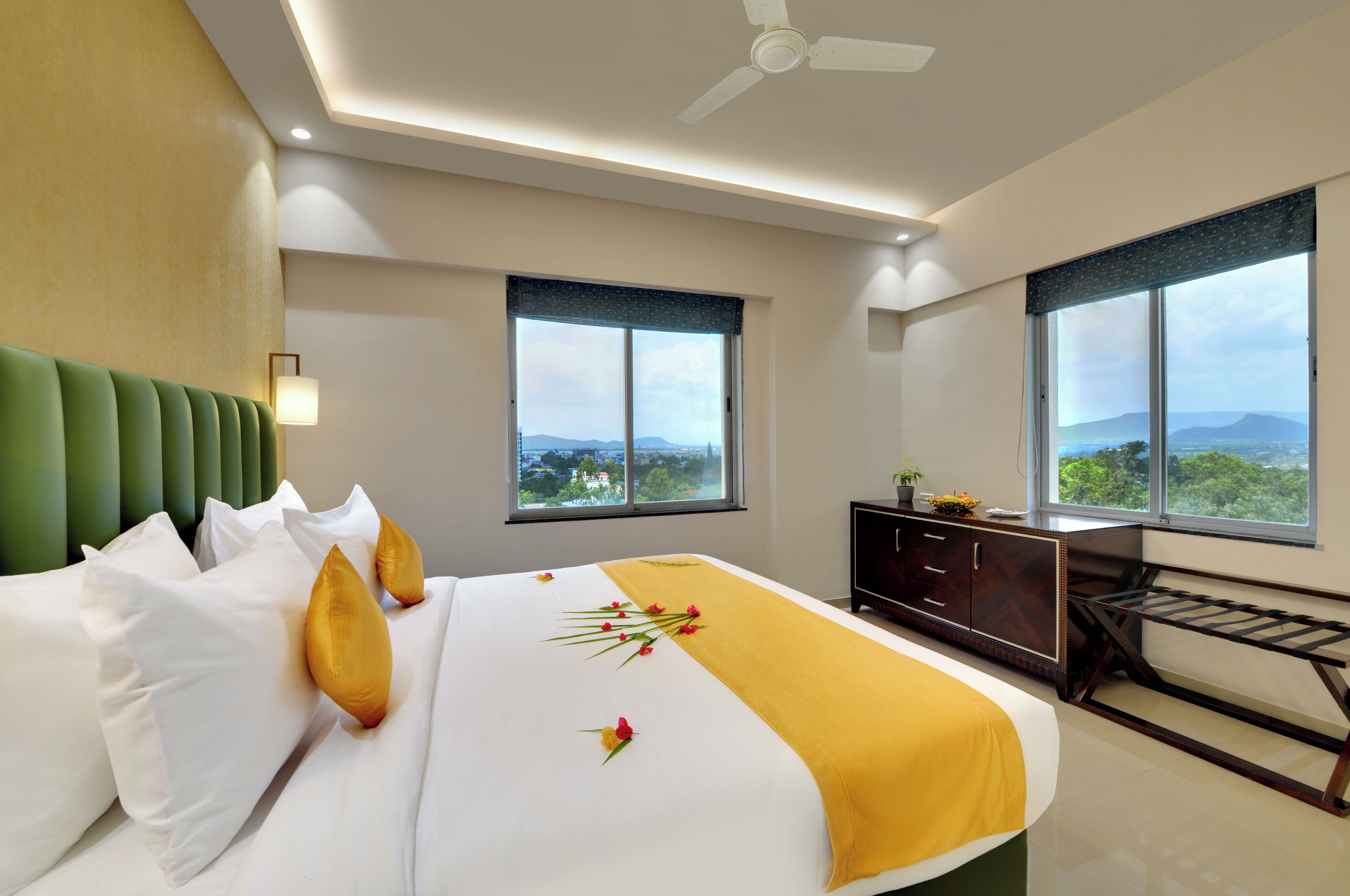 Quarto de hotel moderno com uma cama grande, cabeceira verde, cômoda de madeira e vista para o exterior por duas janelas.
