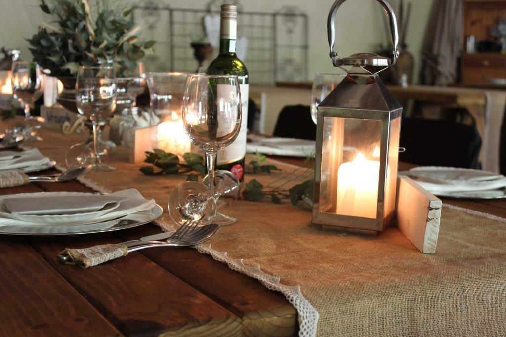 Mesa de jantar posta com taças de vinho, pratos, talheres e uma vela acesa em ambiente rústico.