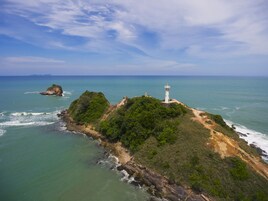 Paisagem costeira com um farol em um afloramento rochoso, rodeado por vegetação e mar.