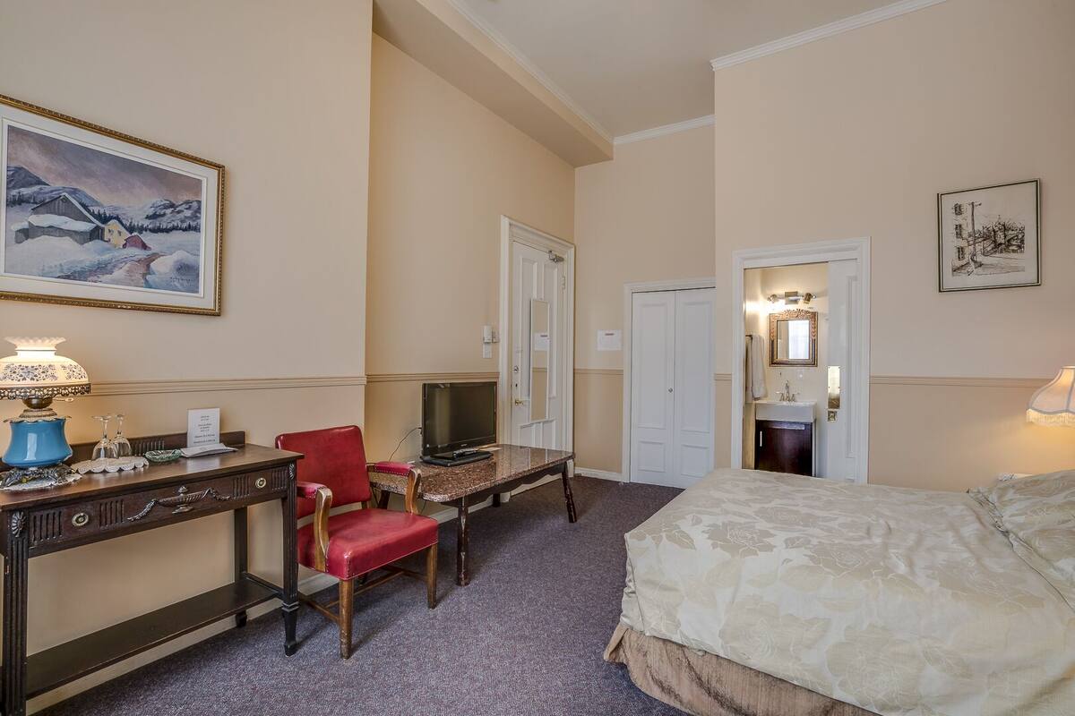 Quarto de hotel com uma cama, uma escrivaninha com computador, uma cadeira vermelha e um quadro na parede.