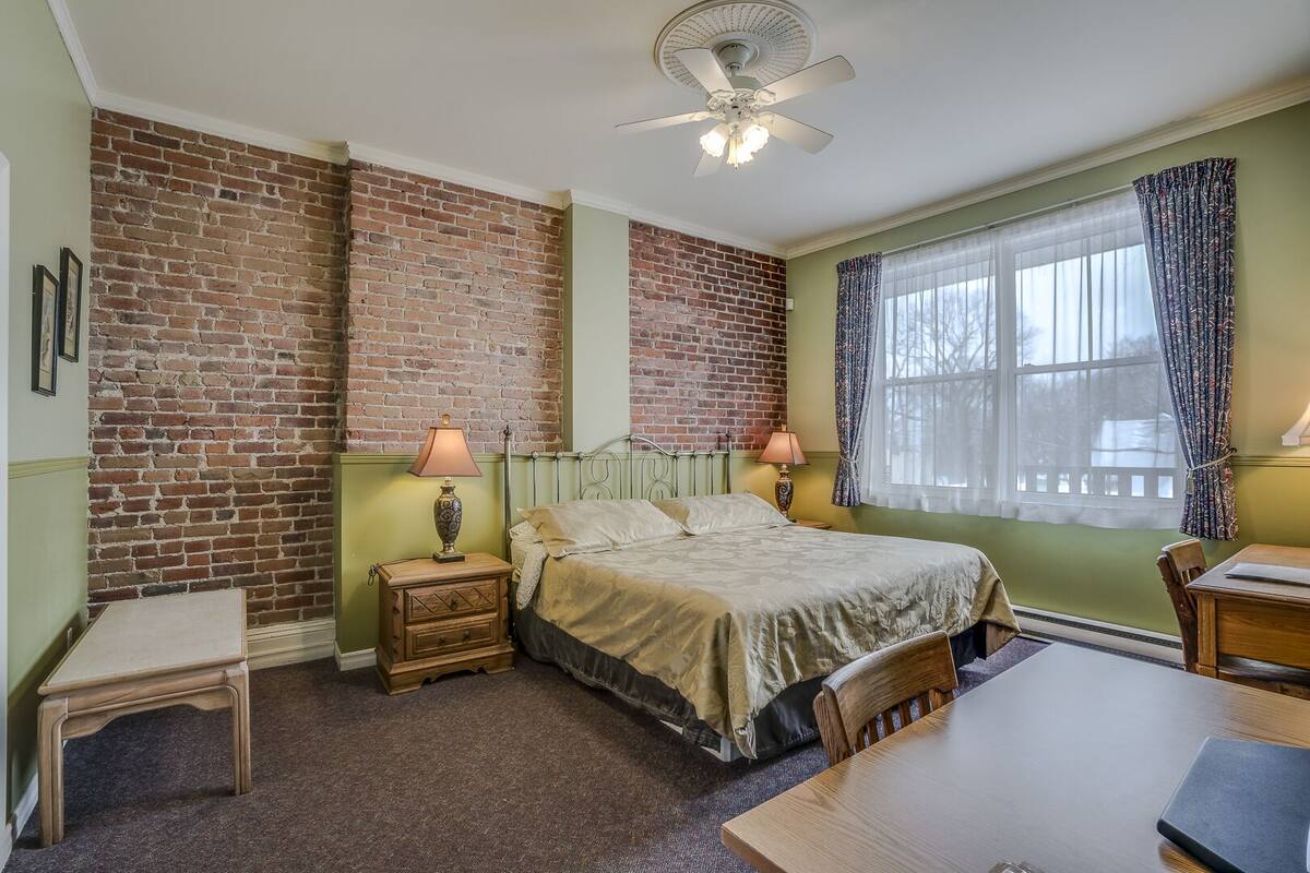 Quarto com parede de tijolos, uma cama com duas mesas de cabeceira, um ventilador de teto e uma janela com cortinas.