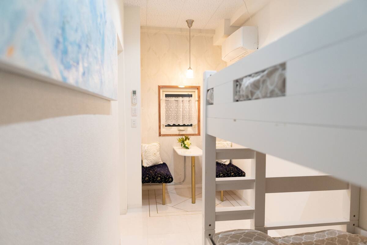 Quarto com beliche, uma pequena mesa e cadeira, janela com cortinas e um quadro na parede.