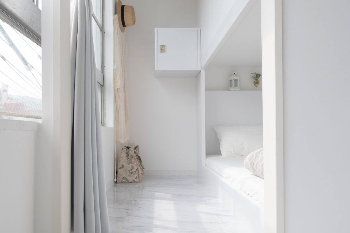 Um corredor estreito leva a um quarto branco com uma cama, um armário pequeno e um chapéu pendurado.