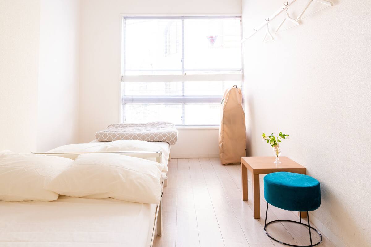 Quarto pequeno e luminoso com duas camas, uma mesinha e uma janela com persianas.