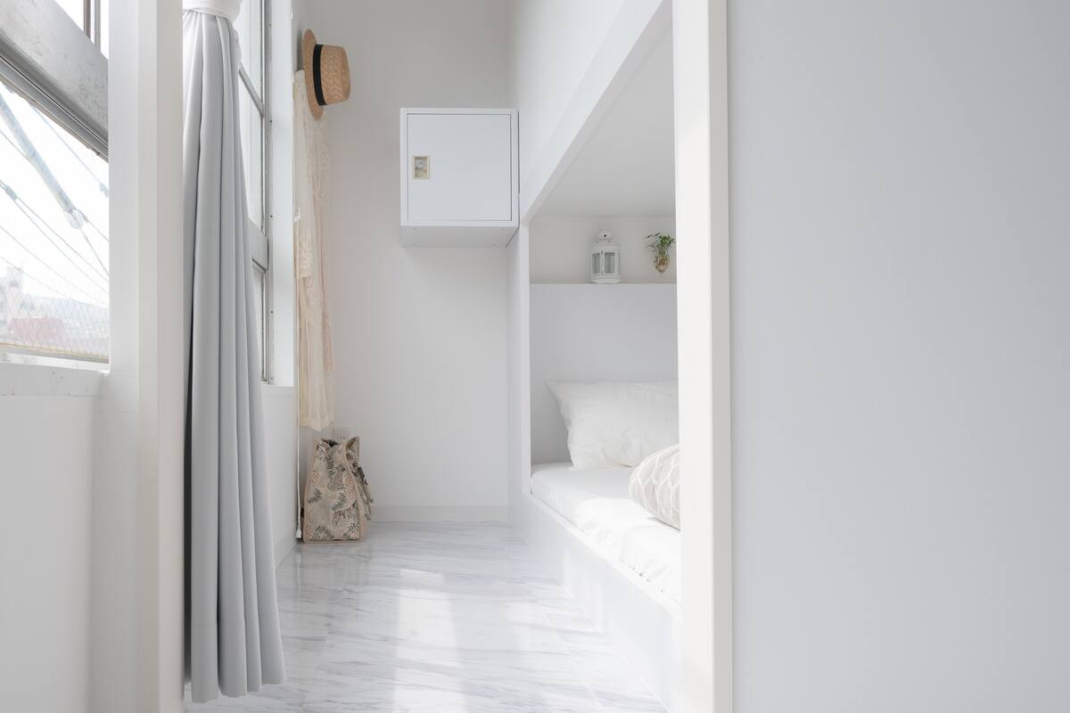 Quarto minimalista com uma cama, um pequeno armário e uma janela com cortinas.