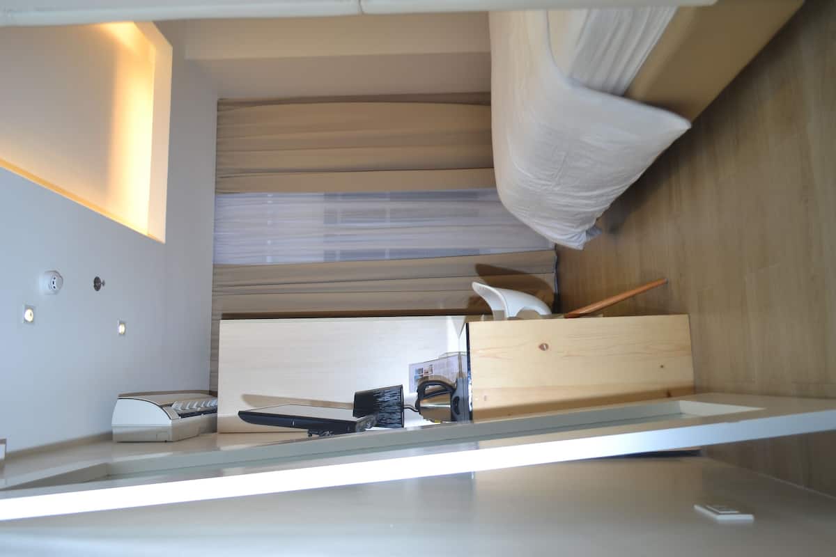 Quarto compacto e moderno, com design minimalista, que inclui uma escrivaninha embutida, um pequeno espaço de estar e uma janela com cortinas leves.