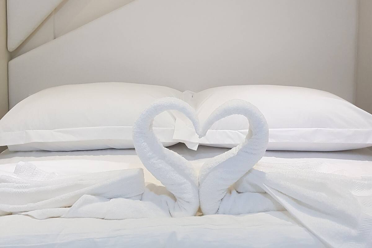 Cama bem arrumada com lençóis e travesseiros brancos, arranjo de toalhas em formato de coração e um design geométrico na parede.