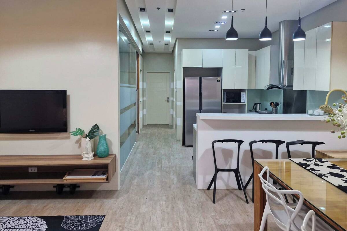 Uma cozinha moderna com uma ilha central, sala de jantar com mesa e cadeiras, e uma TV fixada na parede.