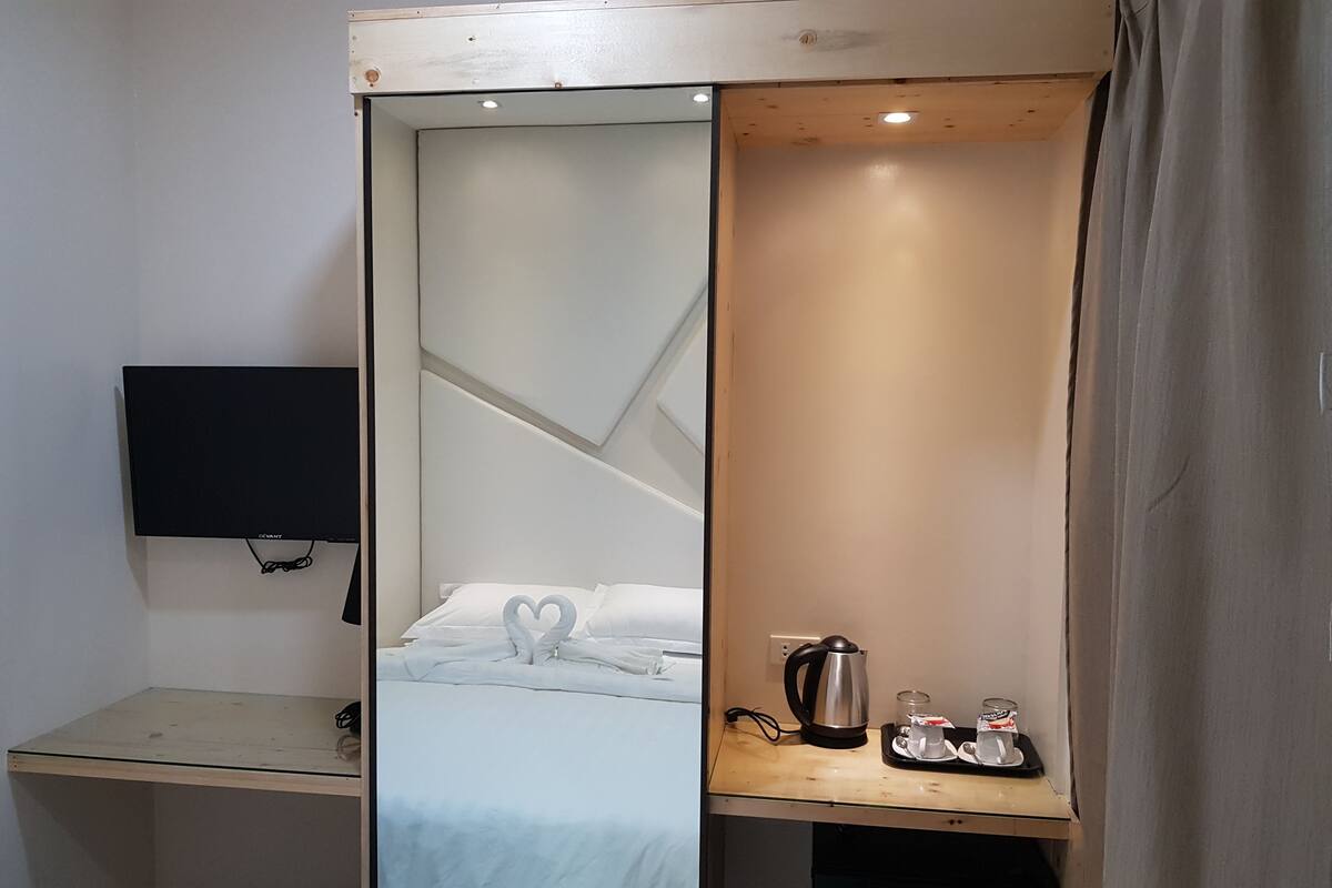 Quarto de hotel com cama, mesa de cabeceira com chaleira e chávenas, espelho e televisão sobre um móvel.