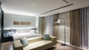 Quarto de hotel moderno com uma cama grande, um sofá, um abajur e vista para a cidade através das cortinas.
